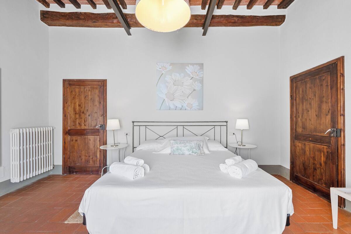 Quarto com uma cama, duas mesas de cabeceira, radiador, portas de madeira e luminária no teto.