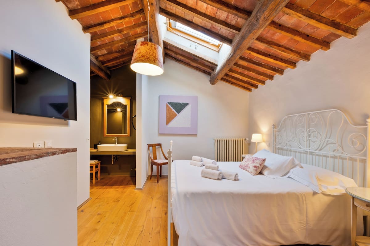 Quarto com cama, televisão, janela, radiador e teto de madeira.