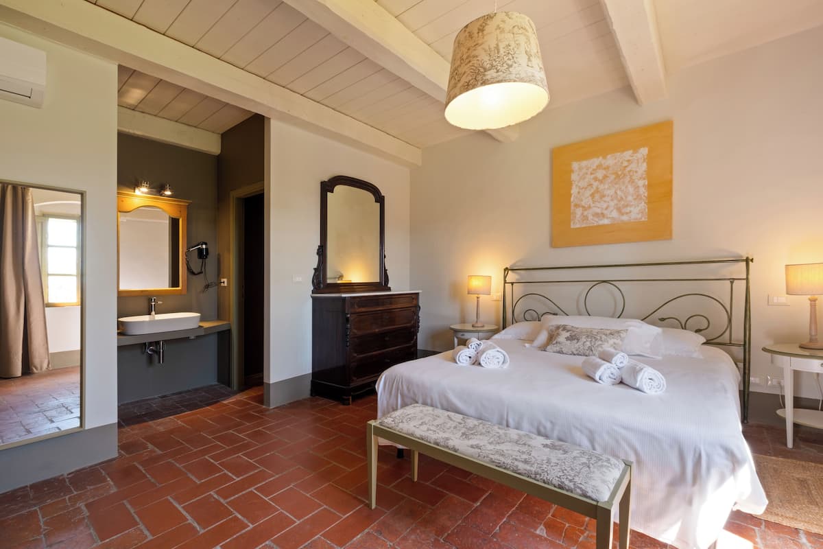 Quarto com cama, mesas de cabeceira, um banquinho, cômoda, espelho e banheiro com pia e espelho.