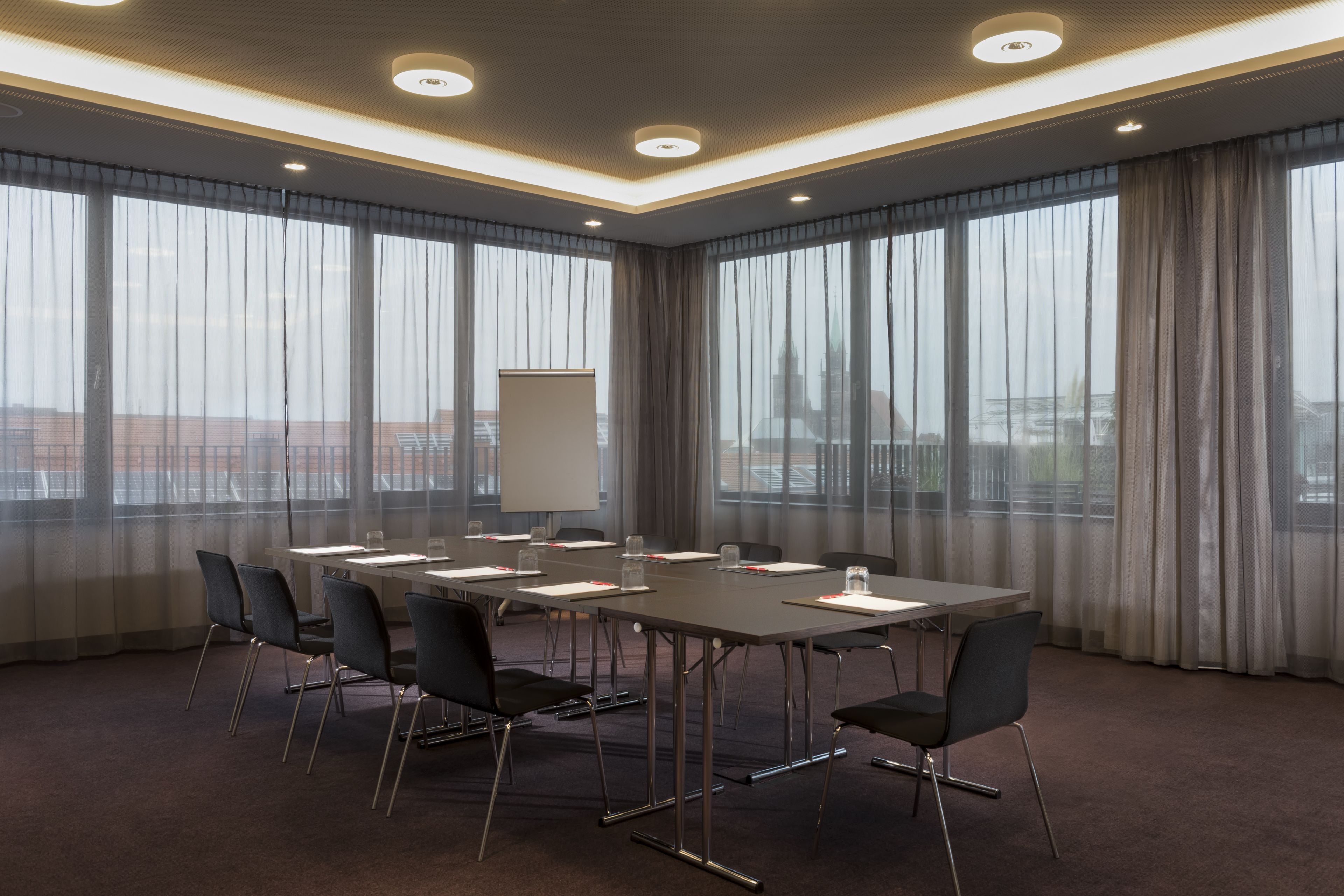 Uma sala de conferência com uma mesa comprida, cadeiras, um quadro branco e janelas amplas com cortinas.