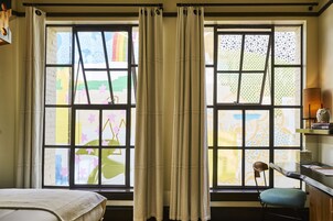 Quarto com cama, escrivaninha e duas janelas com cortinas.