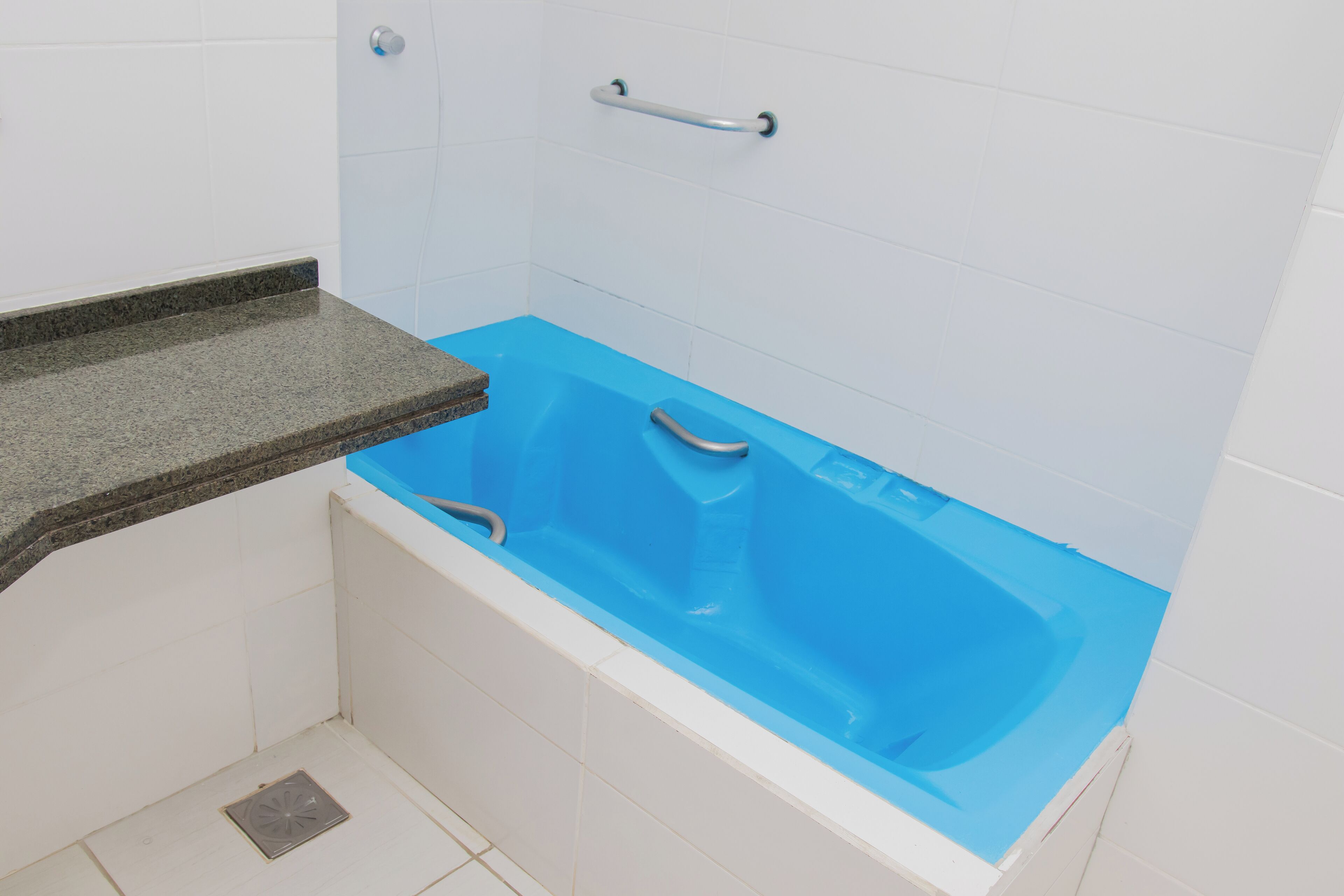 Casa de banho com banheira azul, azulejos brancos, bancada de granito e barra de apoio.