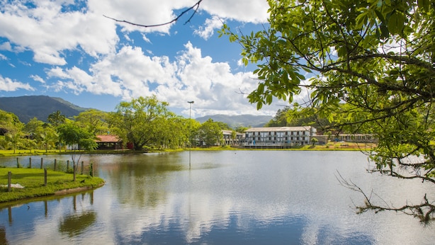 Um lago calmo com o reflexo do céu e vegetação ao redor, um edifício com telhado vermelho e montanhas no fundo.