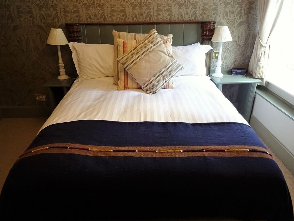 Cama bem arrumada com travesseiro listrado e colcha azul escura. Duas mesas de cabeceira com luminárias e um relógio. O quarto tem papel de parede estampado e uma janela com cortinas.