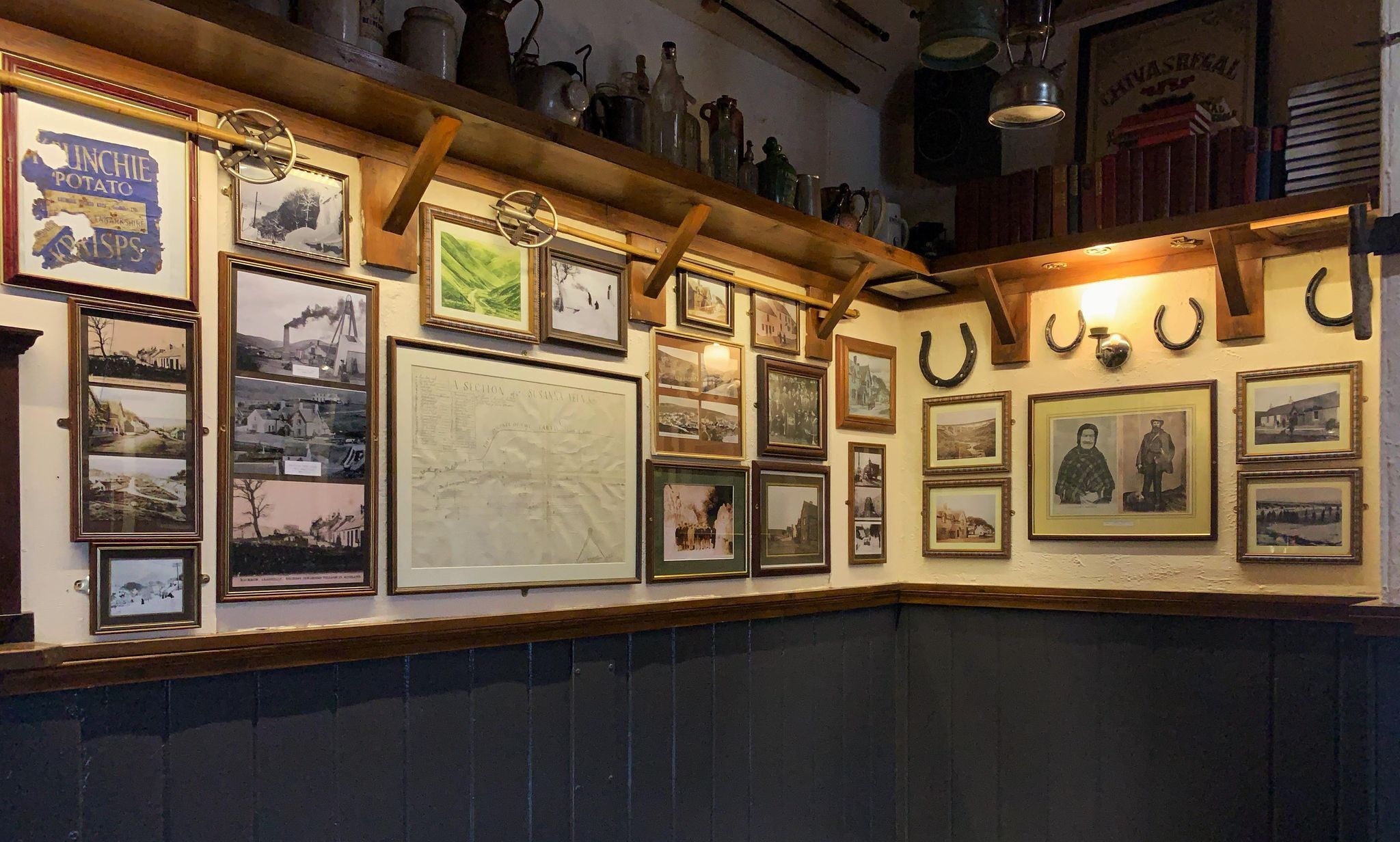 Paredes decoradas com várias fotografias históricas e documentos emoldurados, vigas de madeira e uma estante com diferentes garrafas e recipientes.