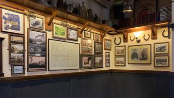 Paredes decoradas com várias fotografias históricas e documentos emoldurados, vigas de madeira e uma estante com diferentes garrafas e recipientes.