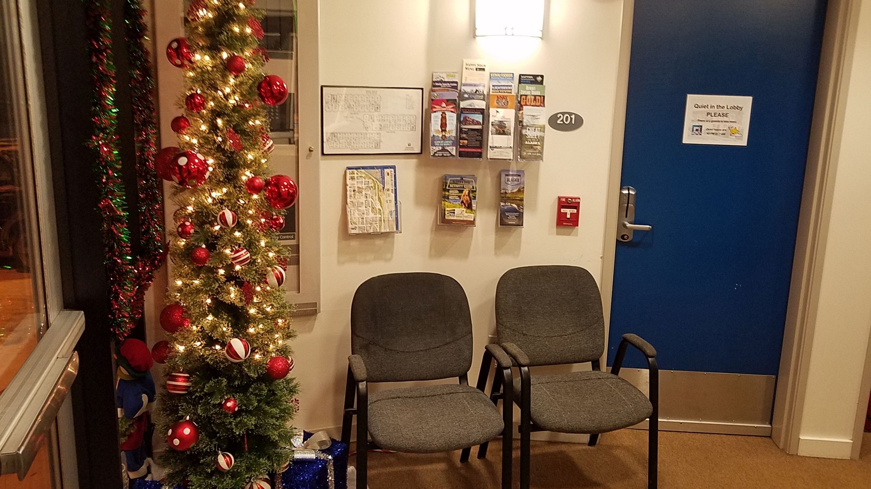 Um quarto com uma árvore de Natal, duas cadeiras cinzentas, uma porta azul e diversos cartazes informativos na parede.