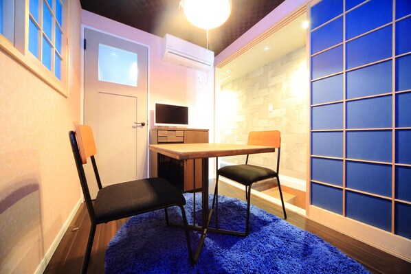Quarto com carpete azul, mesa de madeira, duas cadeiras, televisão e parede com padrão quadriculado.