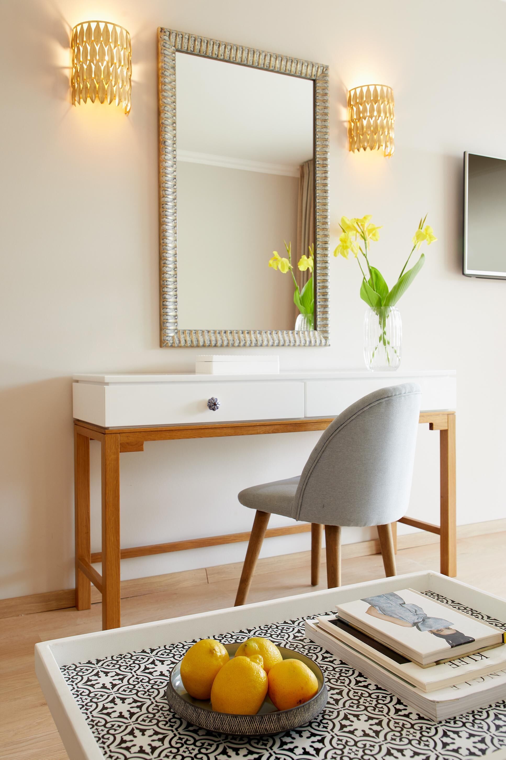 Quarto moderno com escrivaninha de madeira, cadeira cinza, espelho com moldura decorativa e um vaso com flores amarelas.
