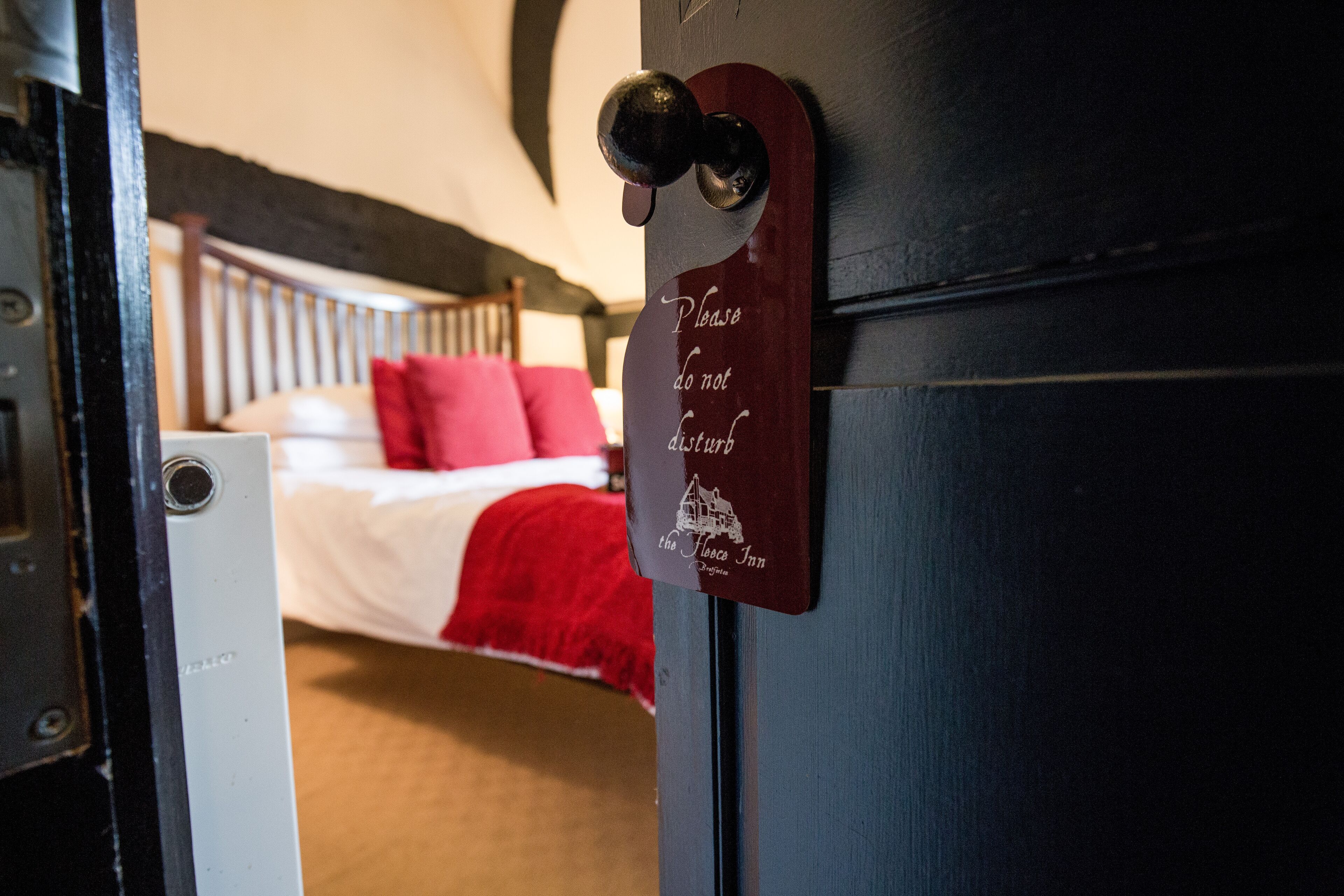 Porta de quarto de hotel com um aviso que diz 'Não perturbe' e uma cama com manta e almofadas vermelhas.