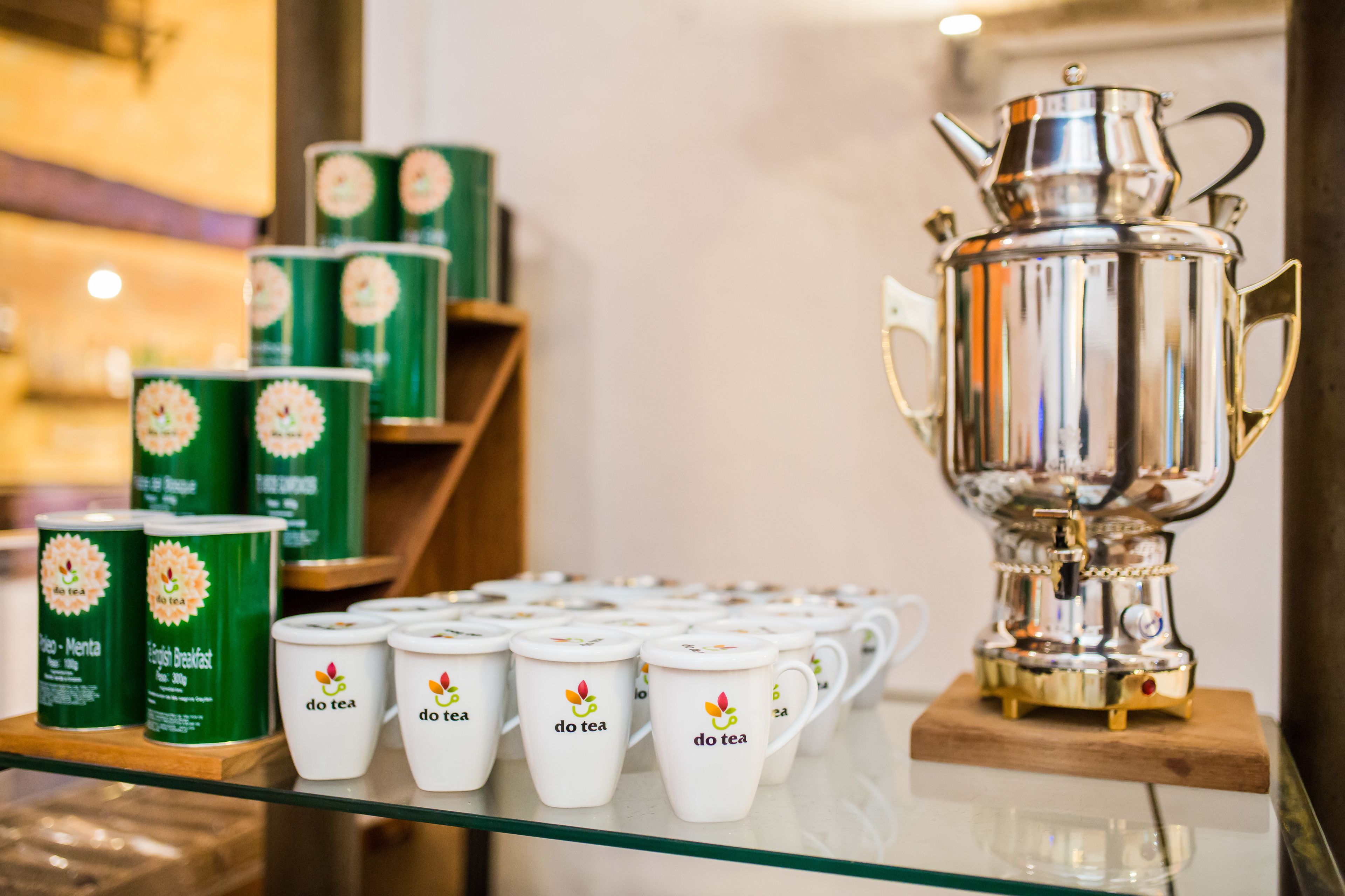 Exposição de produtos de chá, incluindo chávenas brancas com um logótipo, latas de chá verde e um bule de prata numa prateleira de vidro.