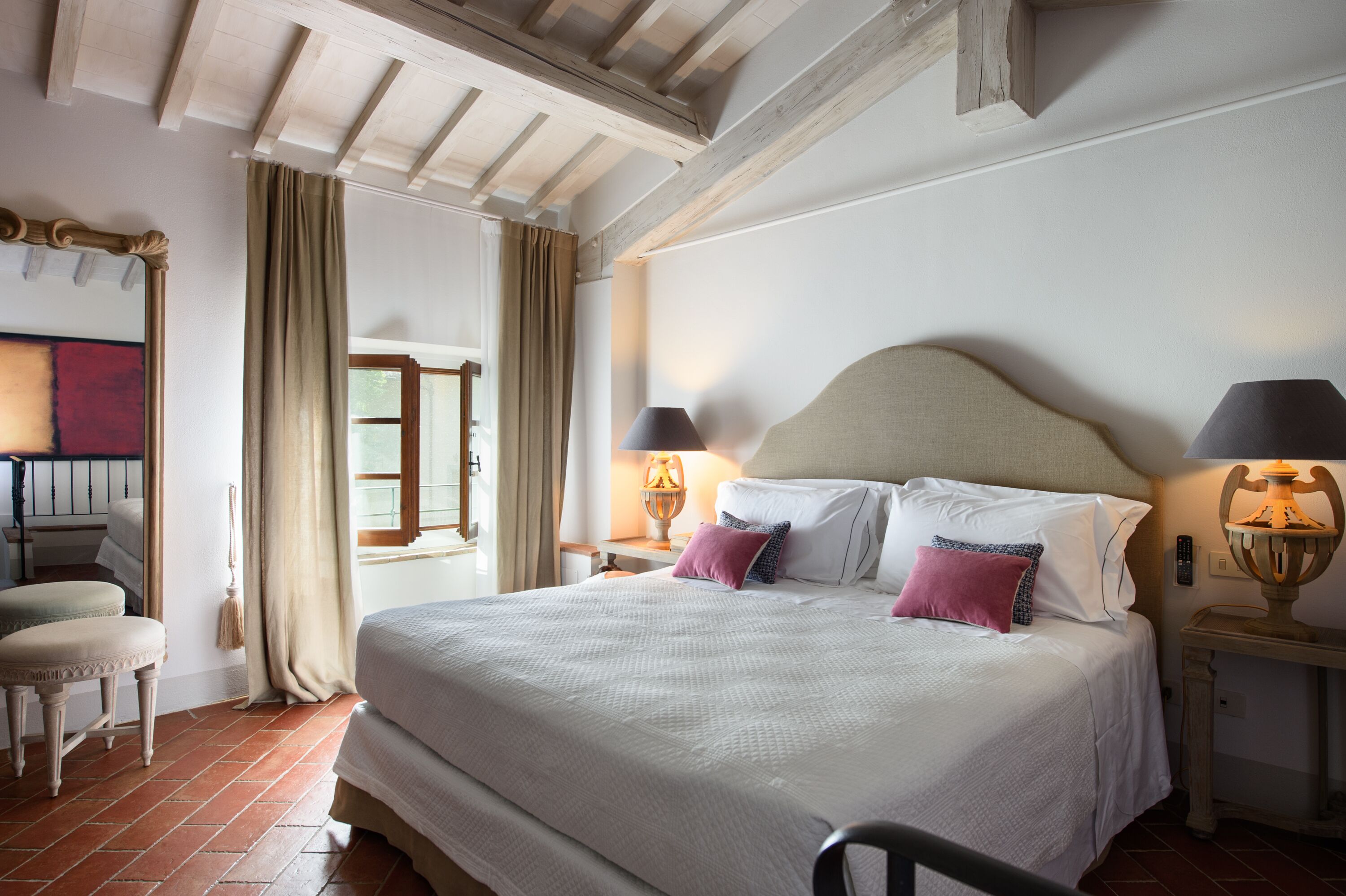 Quarto com cama grande, duas luminárias de cabeceira, mesa de cabeceira, uma cadeira e janela com cortinas.