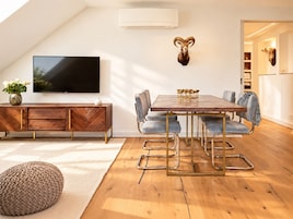 Uma área de refeições moderna com mesa de madeira e cadeiras de metal, televisão montada e um crânio de carneiro decorativo na parede.