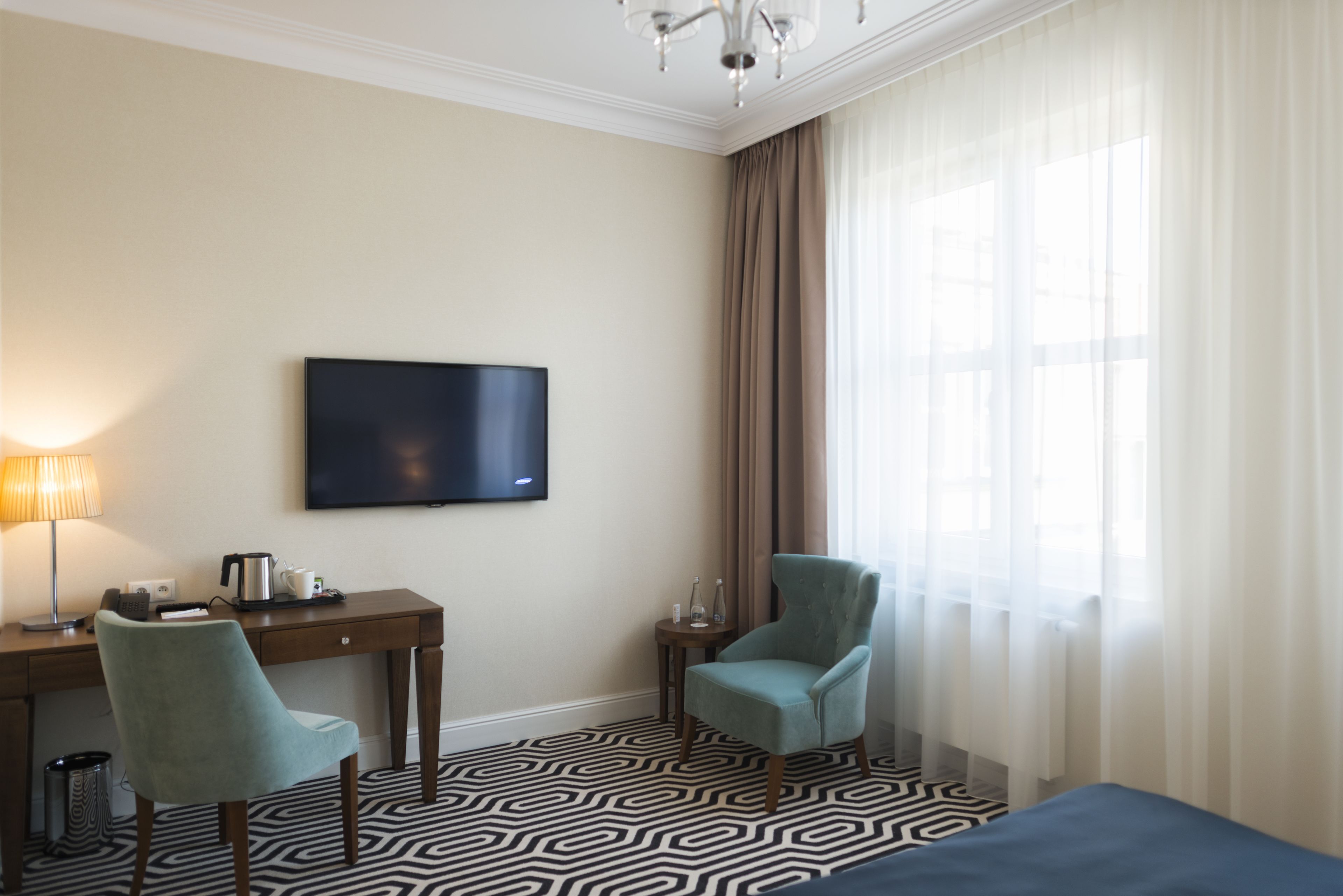 Quarto de hotel com televisão de tela plana na parede, uma escrivaninha com abajur, uma cadeira, uma cama e um tapete estampado.