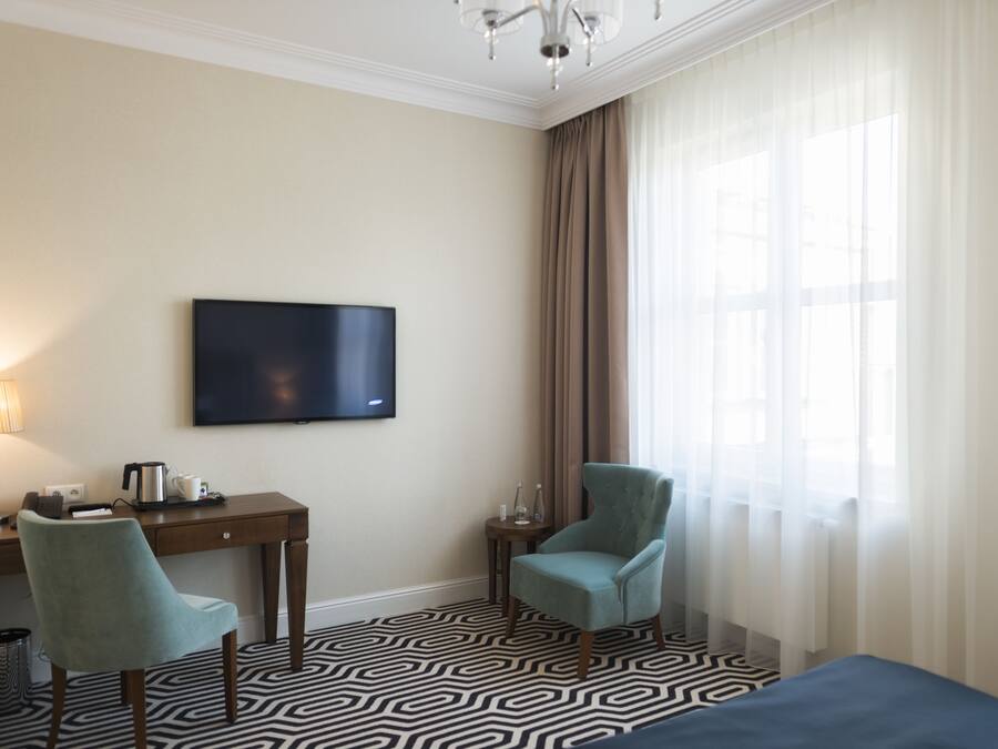 Quarto de hotel com televisão de tela plana na parede, uma escrivaninha com abajur, uma cadeira, uma cama e um tapete estampado.