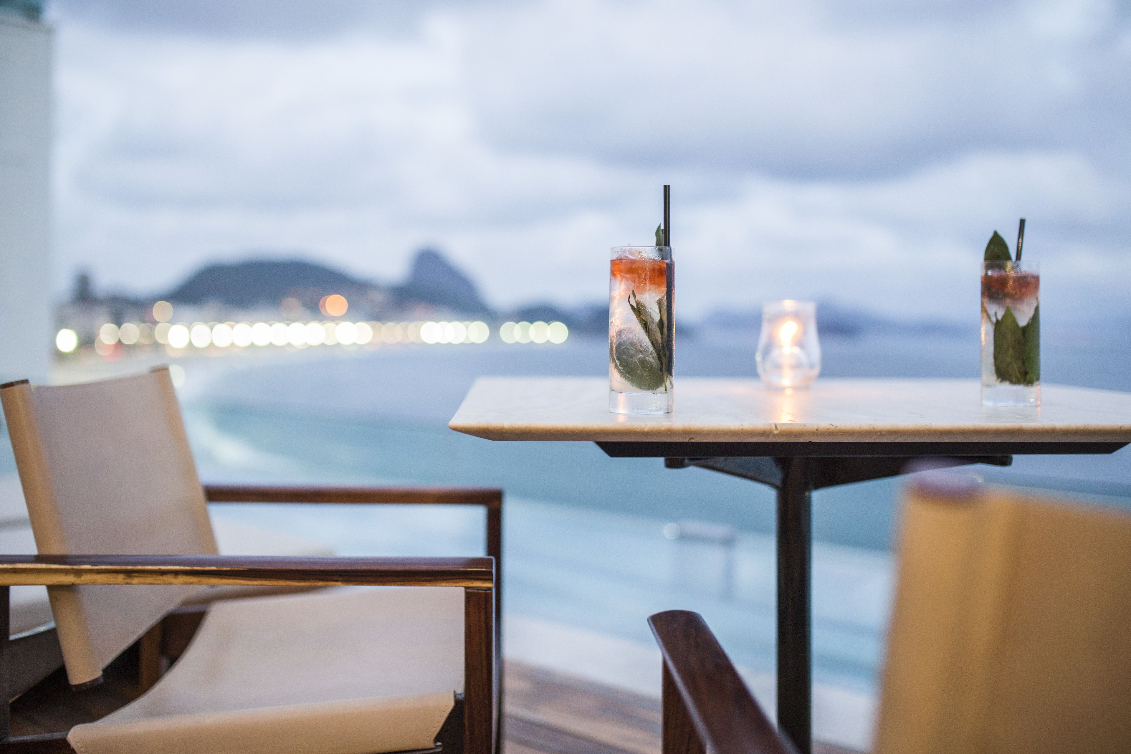Mesa com duas bebidas e uma vela, com vista para um corpo d'água e luzes ao longe.