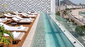 Piscina infinita no terraço, com cadeiras de sol e vista para a praia e o horizonte da cidade.