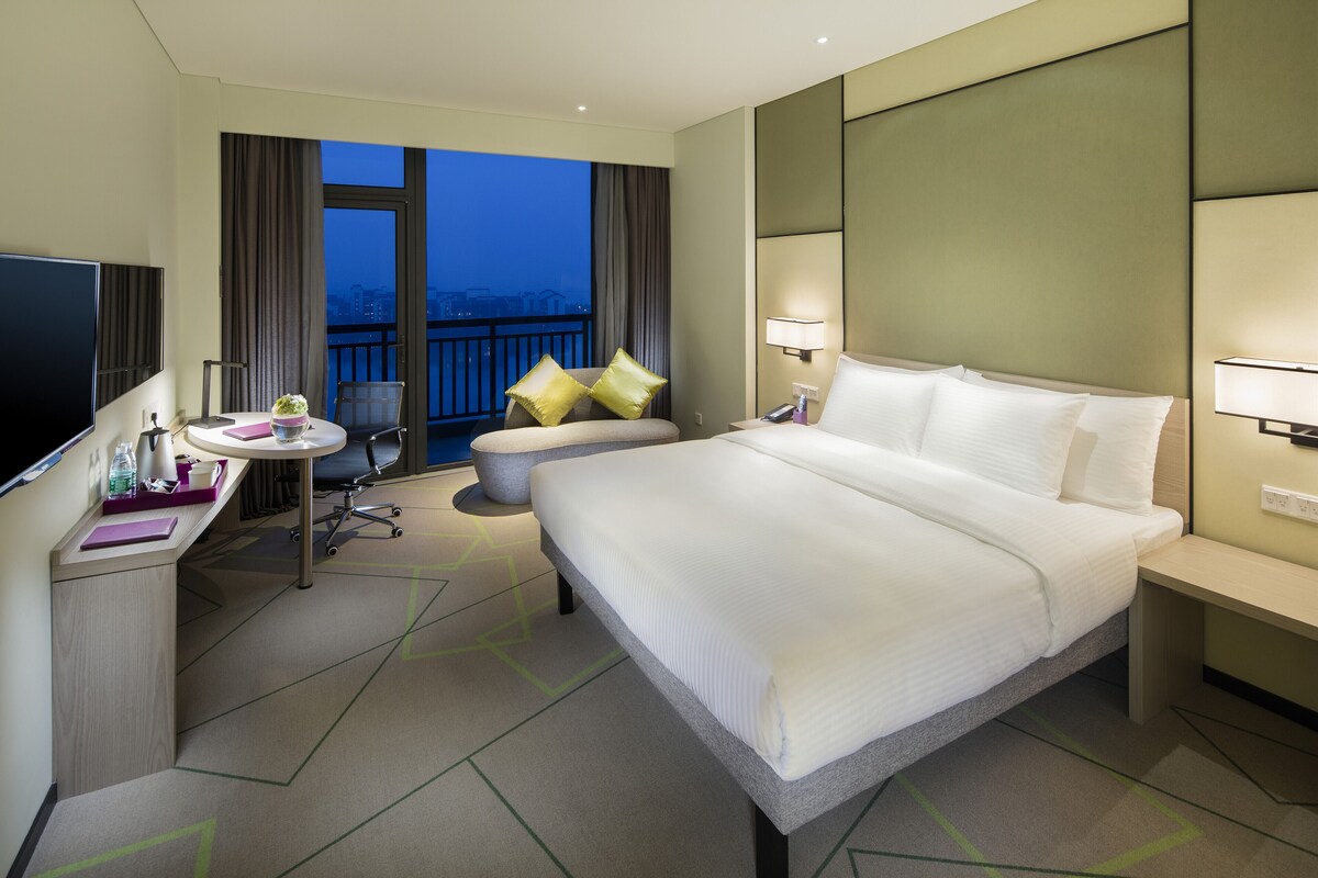 Quarto de hotel com cama grande, escrivaninha com cadeira, televisão e varanda com cortinas.