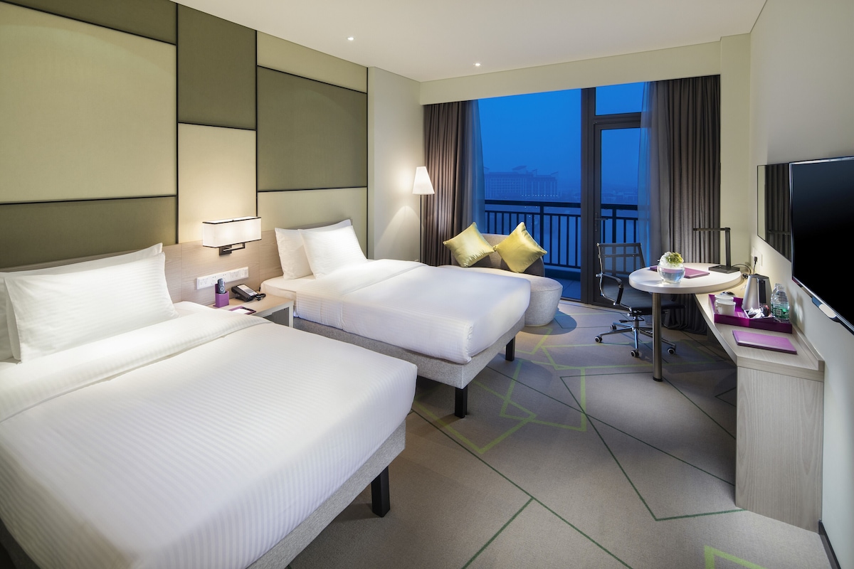 Quarto de hotel com duas camas, uma escrivaninha, uma cadeira, televisão e varanda com vista.