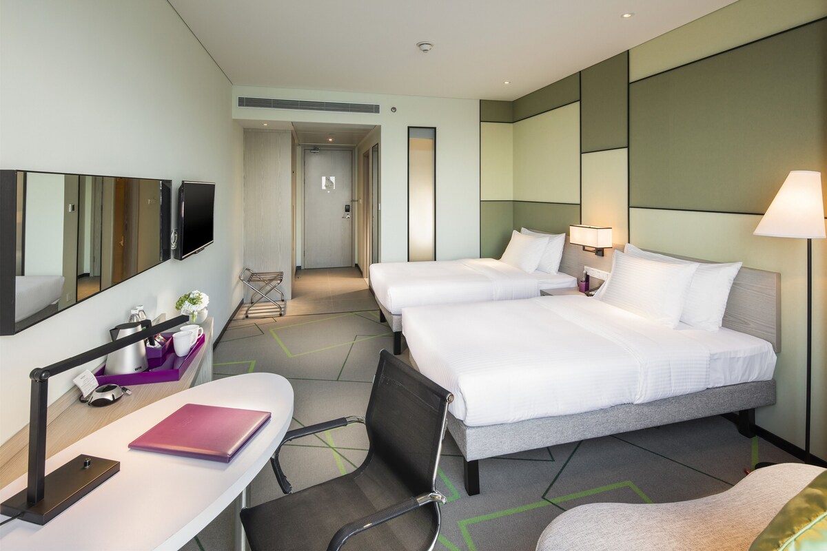 Quarto de hotel com duas camas, uma escrivaninha com cadeira, televisão e banheiro ao fundo.