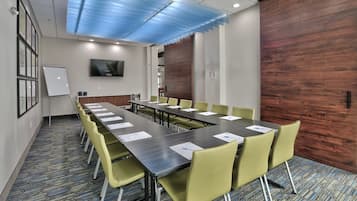 Uma sala de conferências com uma mesa comprida, cadeiras amarelas, um quadro branco e uma televisão na parede.