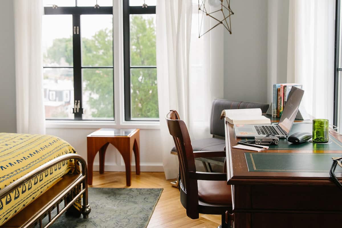Quarto com cama, escrivaninha com laptop e cadeira. Há uma janela com vista para árvores e edifícios.
