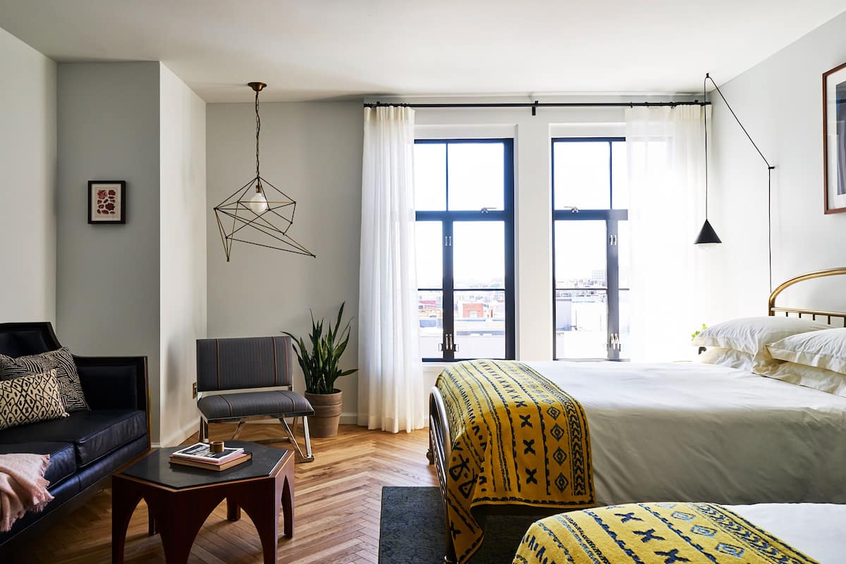 Quarto moderno com sofá preto, uma mesinha, uma cadeira, cama com colcha estampada e uma janela grande com cortinas.