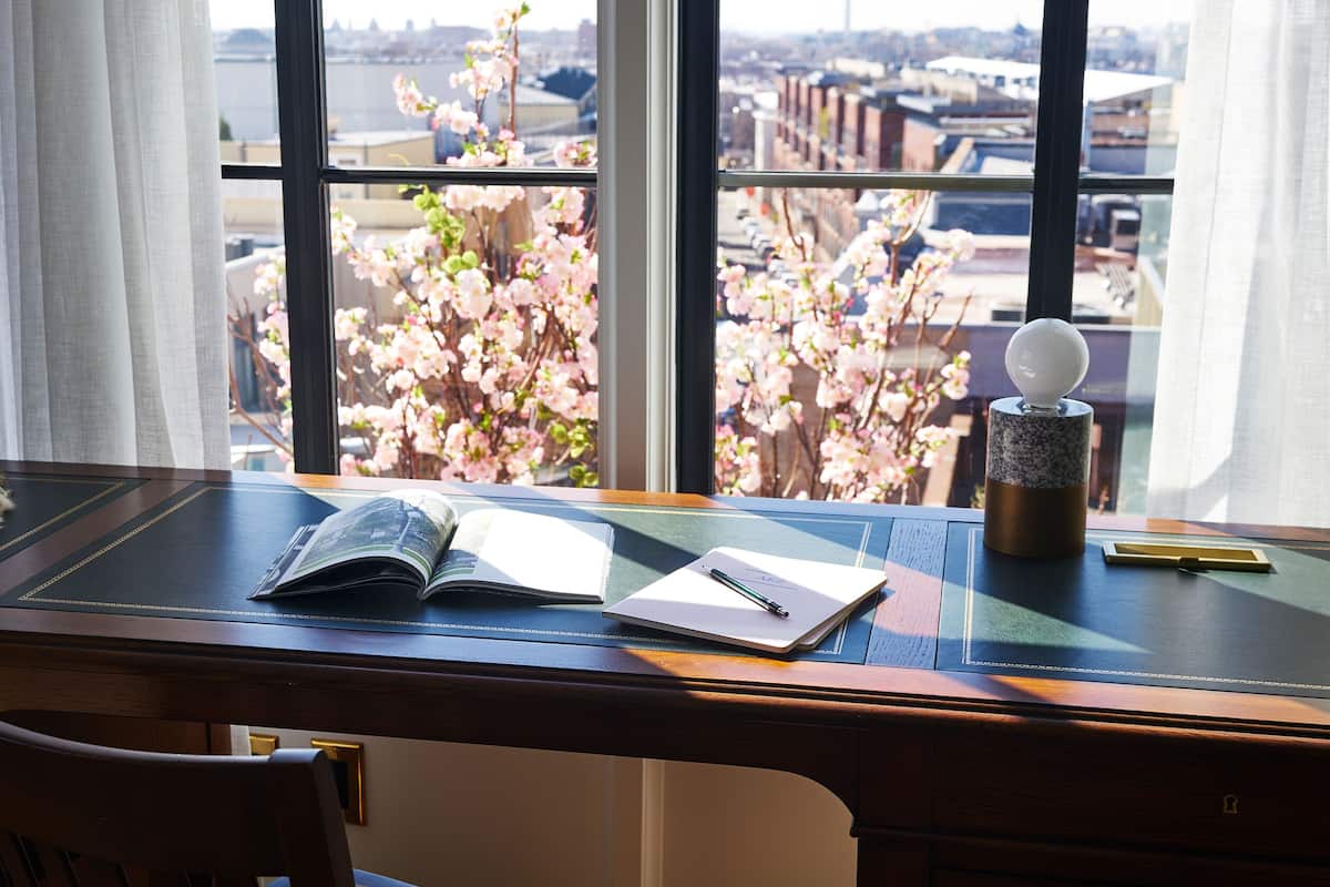 Uma mesa com um livro aberto, um bloco de notas, uma caneta, um objeto decorativo e vista para uma paisagem urbana através de uma janela com flores cor-de-rosa.