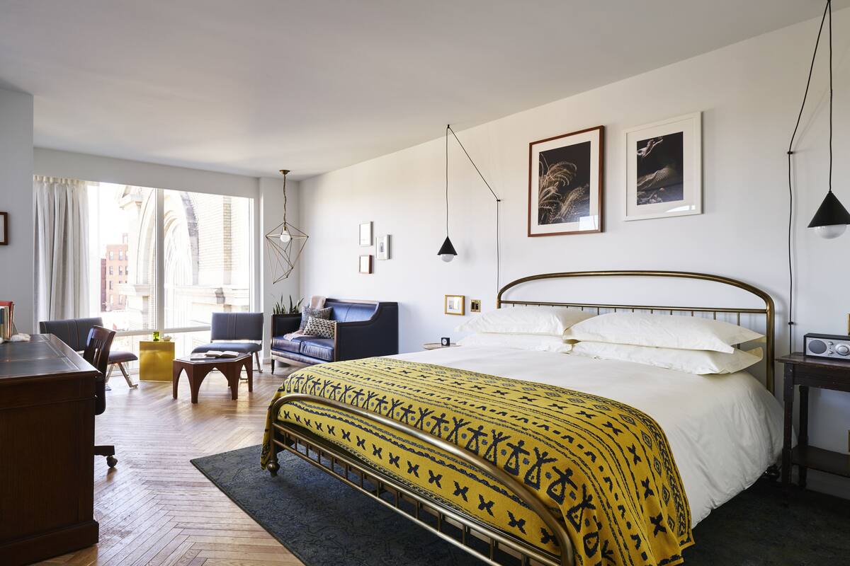 Quarto de hotel moderno com uma cama grande, uma mesa de escritório, uma cadeira e um sofá. Há duas imagens emolduradas na parede e uma janela com cortinas.