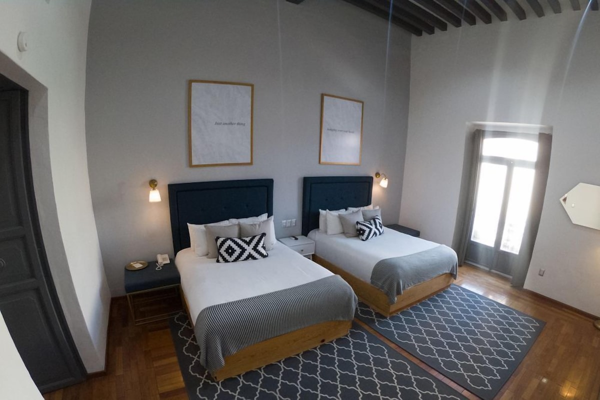 Quarto de hotel com duas camas, uma pequena mesa de cabeceira com abajur, uma porta e uma janela.