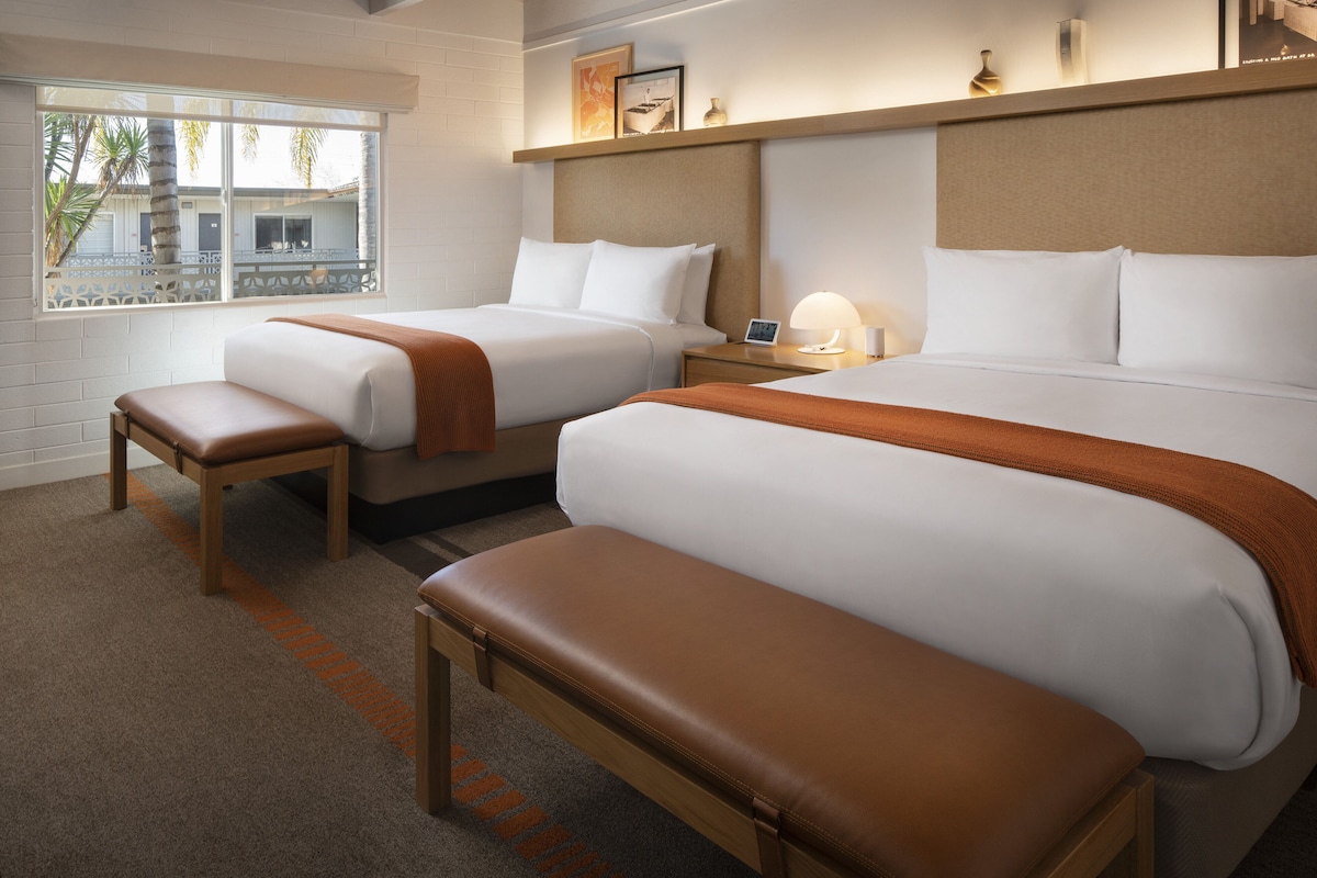 Quarto de hotel com duas camas, um banco, uma mesa de cabeceira com abajur e uma janela com vista para palmeiras.