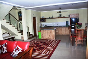 Ampla sala de estar com um sofá vermelho, uma mesa de centro de madeira e um tapete estampado. Uma cozinha com geladeira, pia e armários de madeira é visível ao fundo.