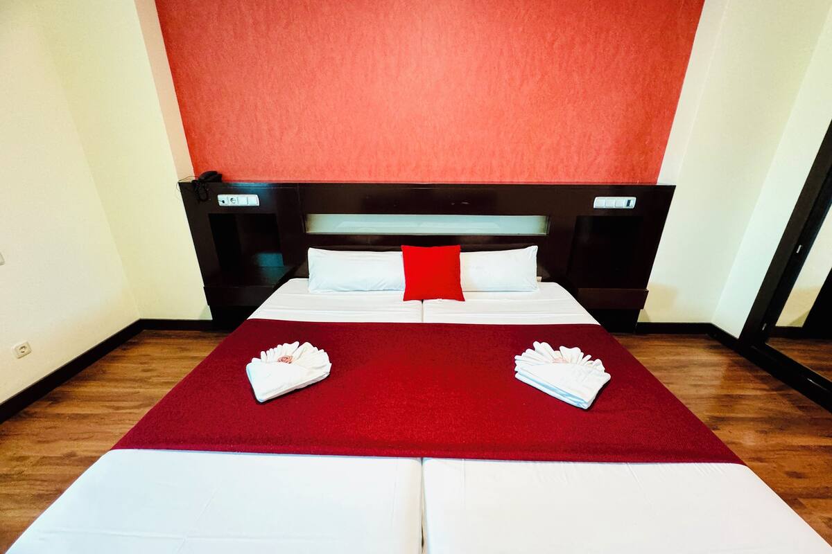 Quarto de hotel com uma cama grande, uma parede de destaque vermelha e duas mesas de cabeceira com luminárias.