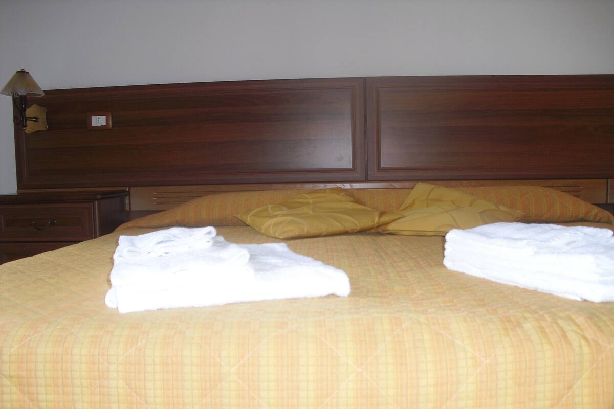Quarto de hotel com uma cama grande, duas almofadas, uma mesa de cabeceira e um abajur.