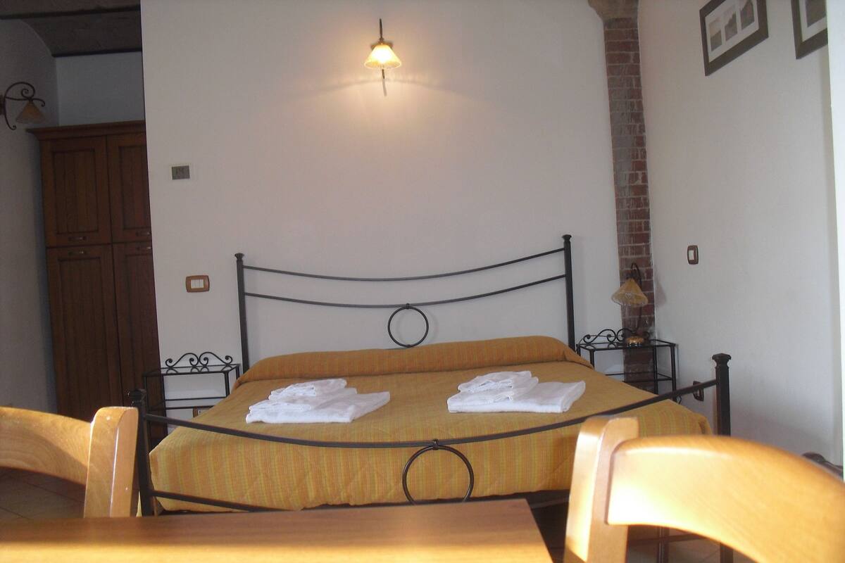 Quarto com cama, cadeiras de madeira, mesa, abajur e luminária na parede.