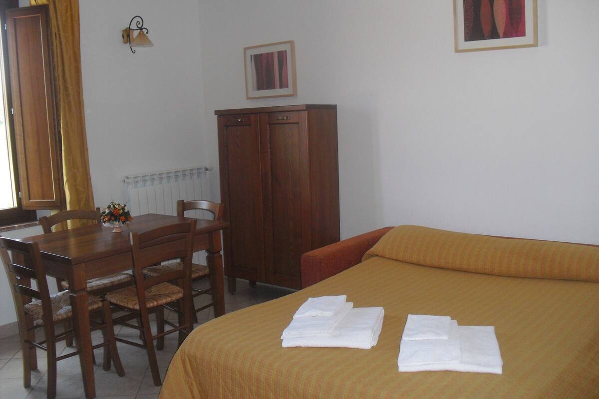 Quarto de hotel com cama, escrivaninha de madeira, cadeiras, armário e radiador.