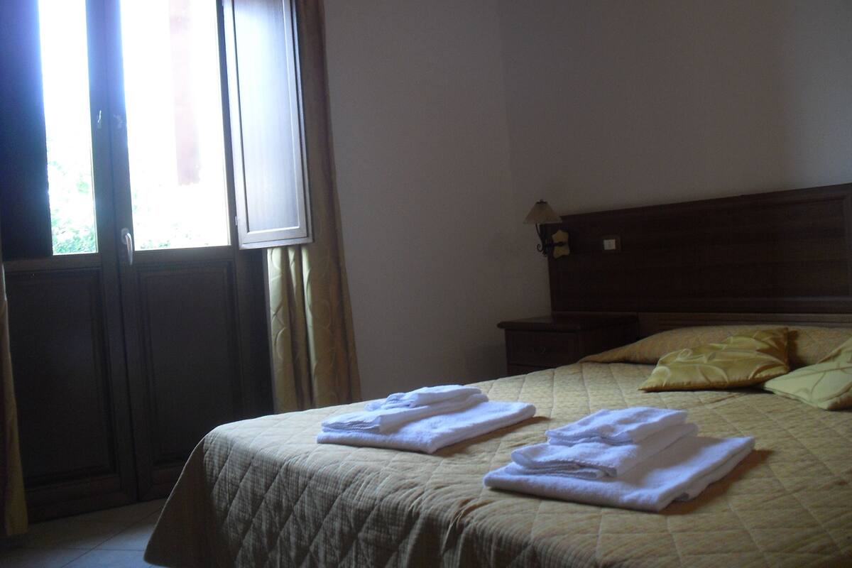 Quarto de hotel com cama, mesa de cabeceira e janela com cortinas.