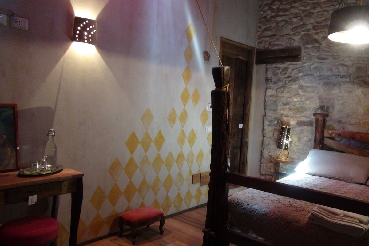 Um quarto com um banco de madeira, um banquinho pequeno, uma mesa com uma garrafa e uma parede com desenho em losangos.