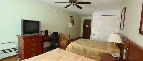 Quarto de hotel com duas camas, uma escrivaninha com computador, televisão, ventoinha de teto e telefone.