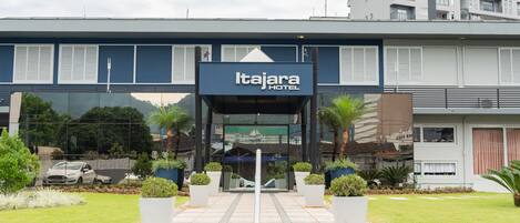 Entrada do Hotel Itajara com design moderno, apresentando fachada de vidro e escadaria que leva à entrada.