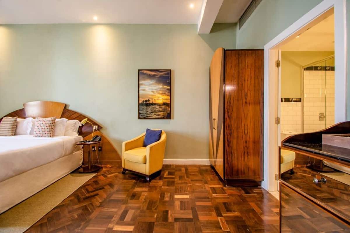 Quarto de hotel com cama, poltrona, escrivaninha, piano e um quadro na parede.