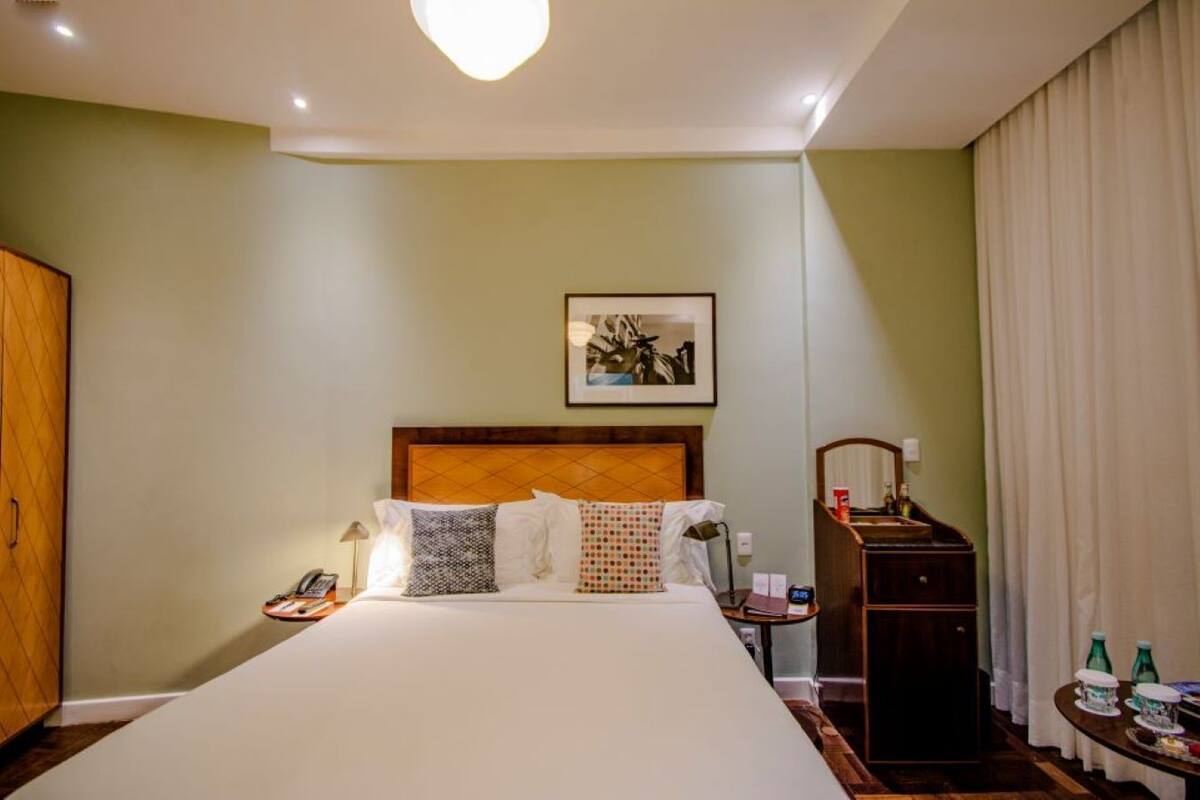 Quarto de hotel com uma cama grande, cabeceira de madeira, mesa de cabeceira com abajur e uma pequena mesa com garrafas e copos.