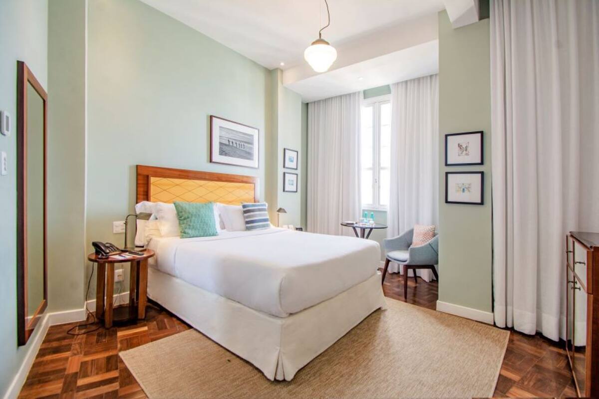 Quarto de hotel com uma cama grande, mesa de cabeceira com telefone, uma cadeira e escrivaninha com abajur.