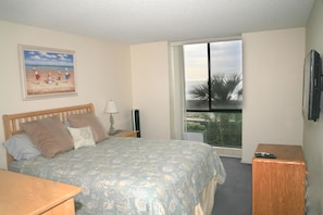 Quarto com cama, mesa de cabeceira de madeira, luminária, televisão e janela com vista para palmeiras.