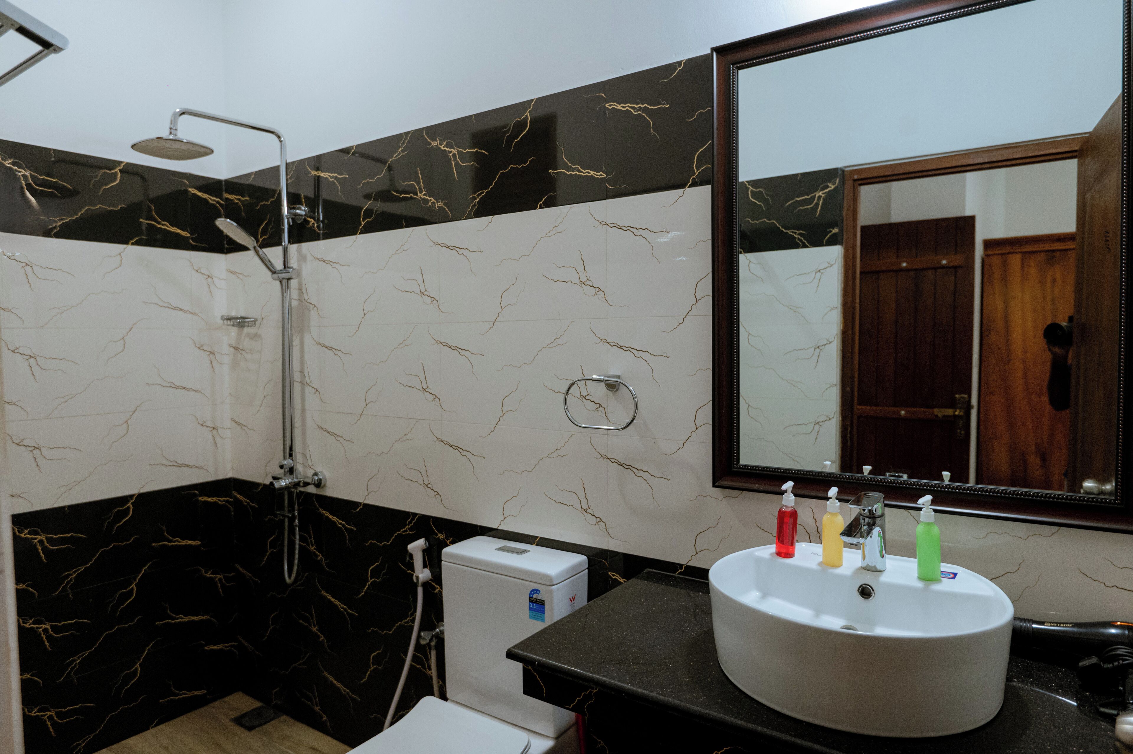 Casa de banho com painel de azulejos de mármore preto e branco, uma pia branca com vários dispensadores de sabonete, um espelho e um chuveiro com chuveiro manual.