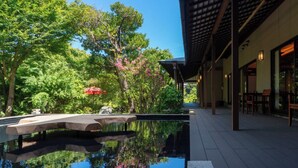 Uma área de piscina ao ar livre tranquila, com um banco de pedra, cercada por vegetação exuberante e um edifício tradicional com colunas de madeira.