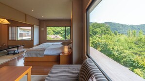 Quarto com uma janela ampla, uma cama, mesa de cabeceira e vista para uma área arborizada.