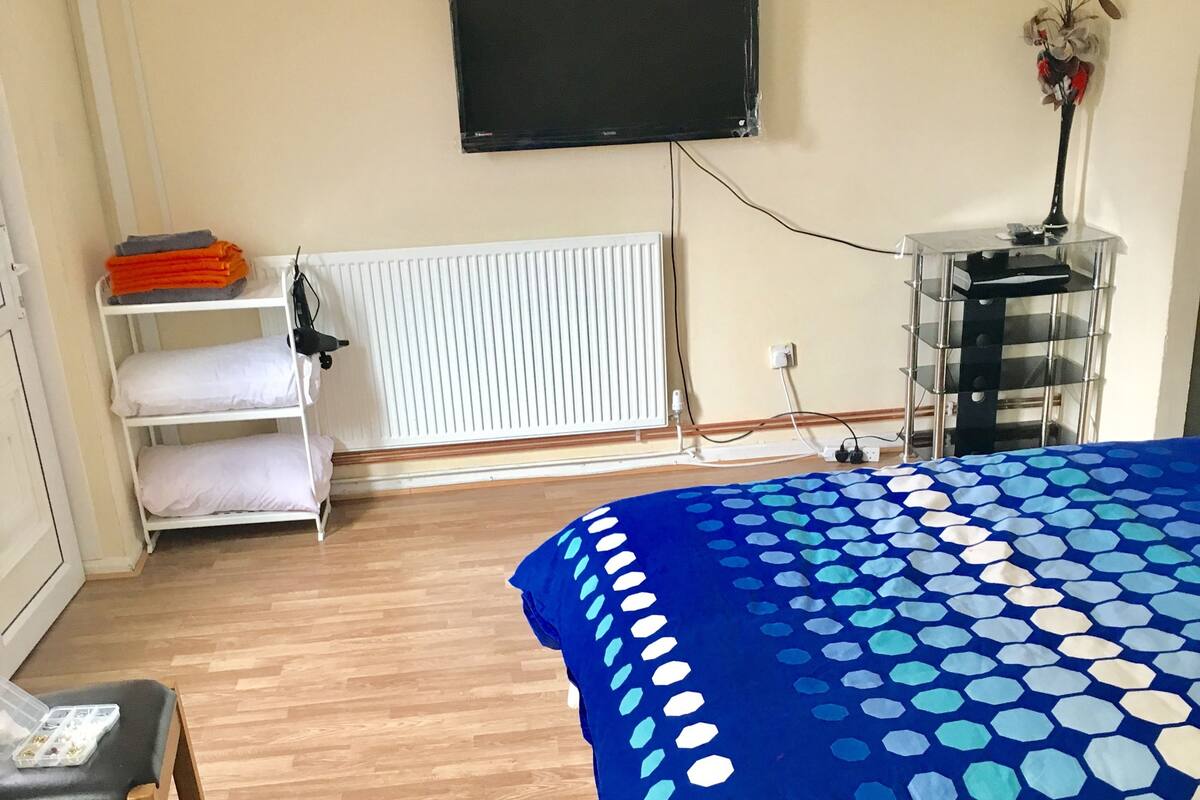 Quarto com cama, cadeira, radiador, prateleira com roupas dobradas e televisão fixada na parede.
