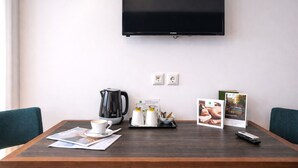 Quarto de hotel com TV de tela plana na parede, mesa de madeira com xícara de café, chaleira, bandeja com condimentos e controle remoto.