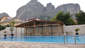 Piscina com área de estar coberta, rodeada por plantas em vasos e um pano de fundo de grandes formações rochosas.